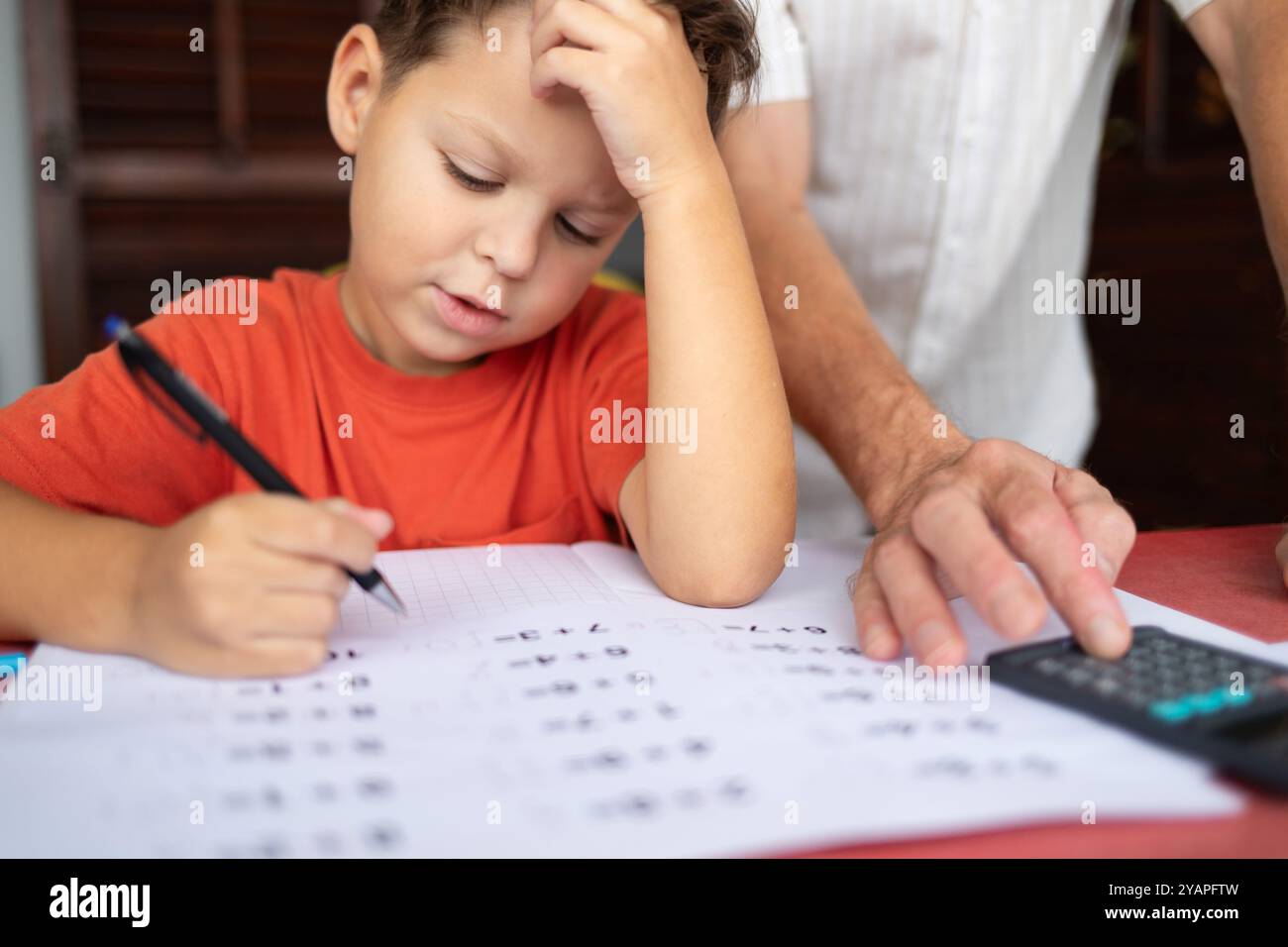 A boy does his math homework while his father stands next to him ...