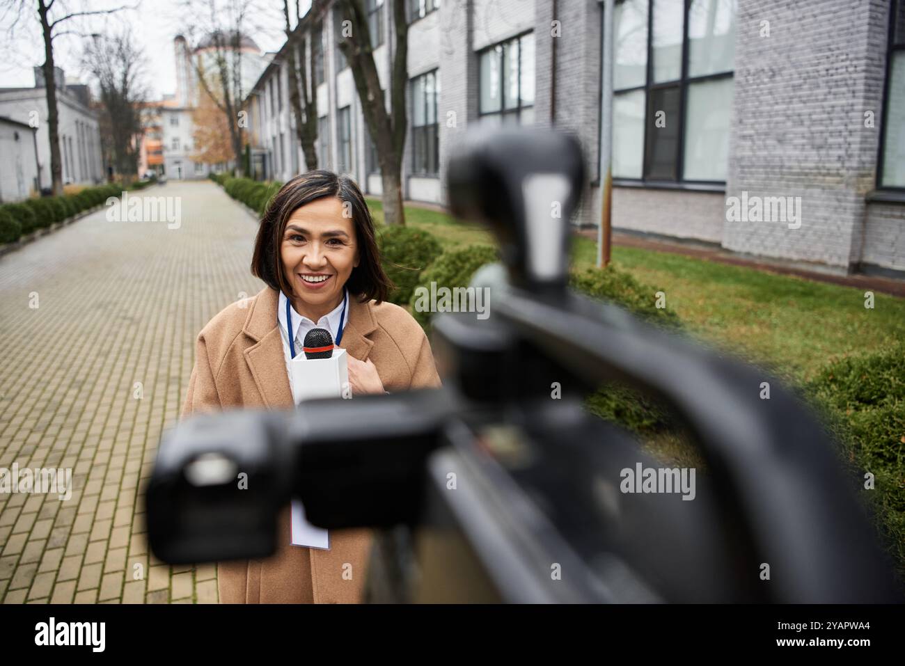 A multiracial female journalist stands outdoors, smiling and reporting ...