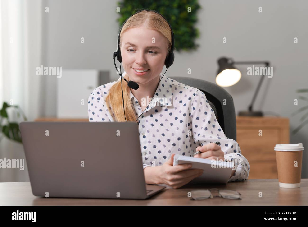 Interpreter in headset taking notes while working with laptop at table ...