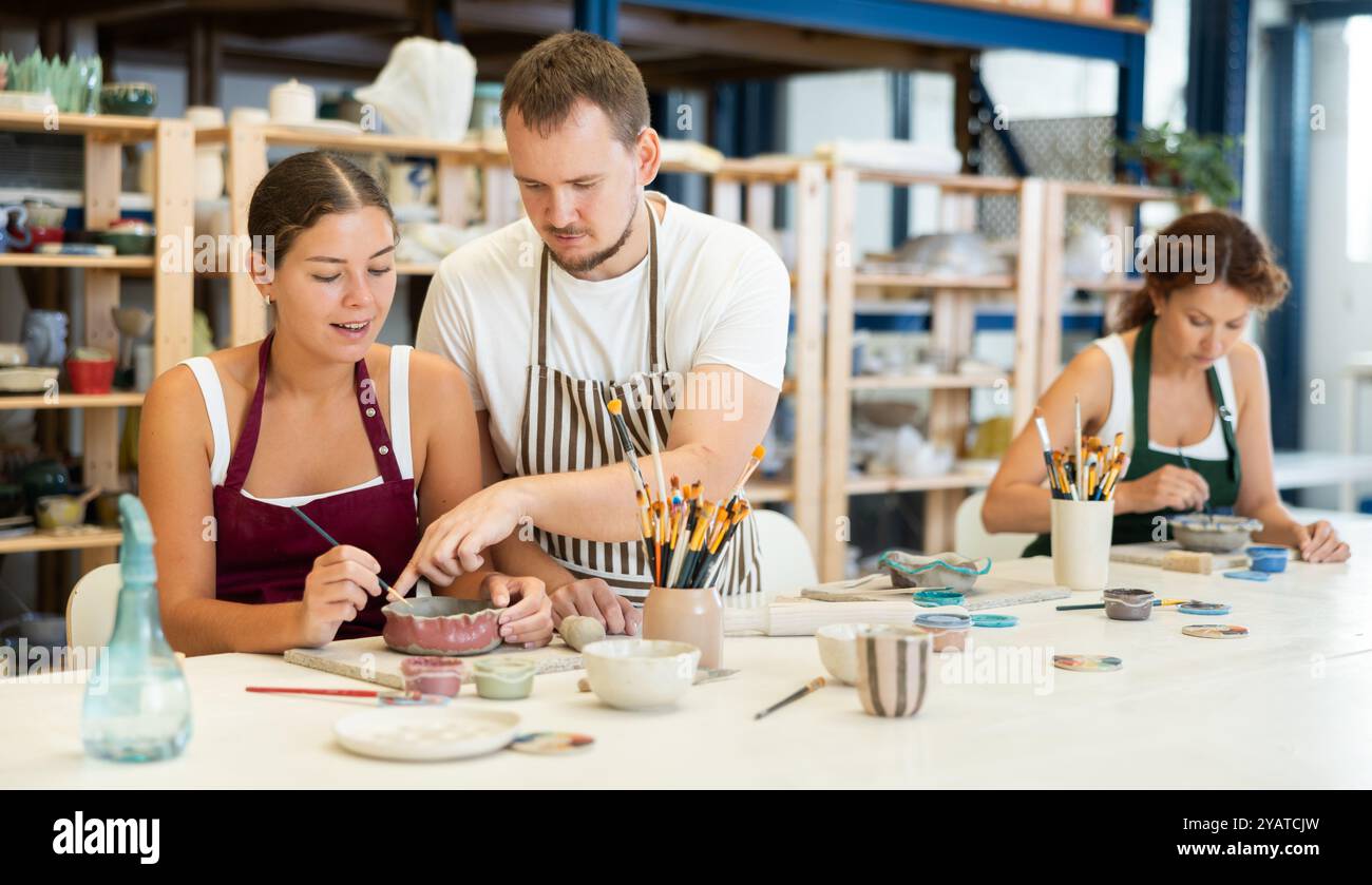 During master class, student paints clay vessel under guidance of male ...