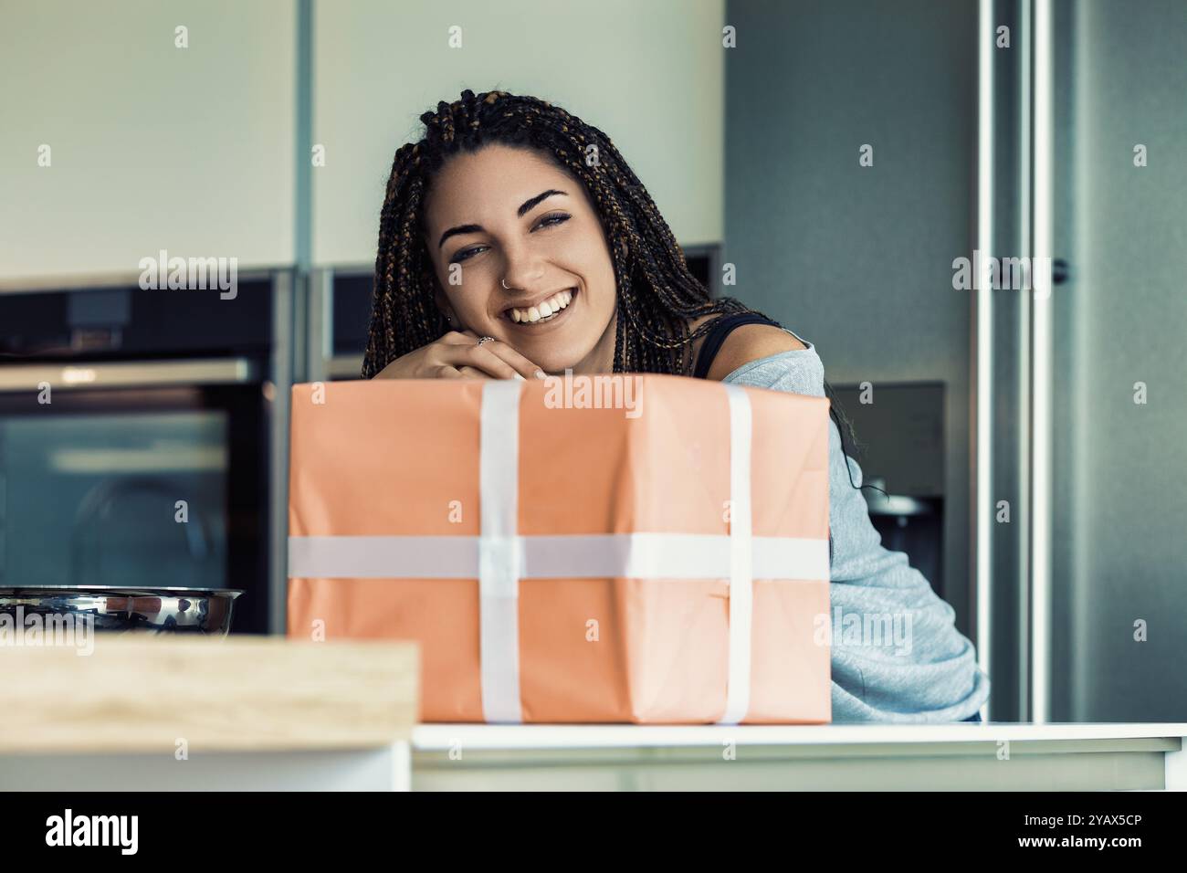 Young woman smiles happily in her cozy kitchen, leaning on a gift box ...