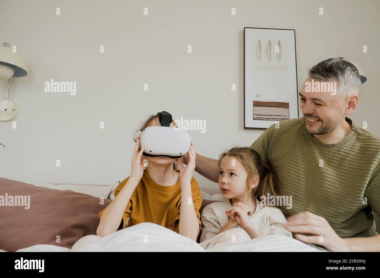 A father sits on a bed, watching his daughter experience a virtual reality world with a VR ...