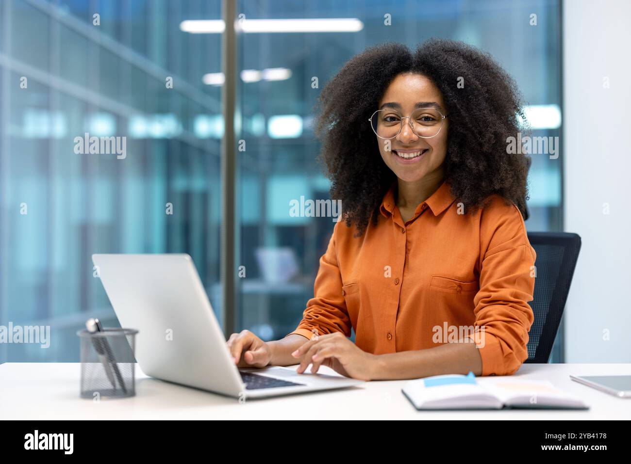 Young professional woman smiling confidently while working on laptop at office desk, showcasing ...