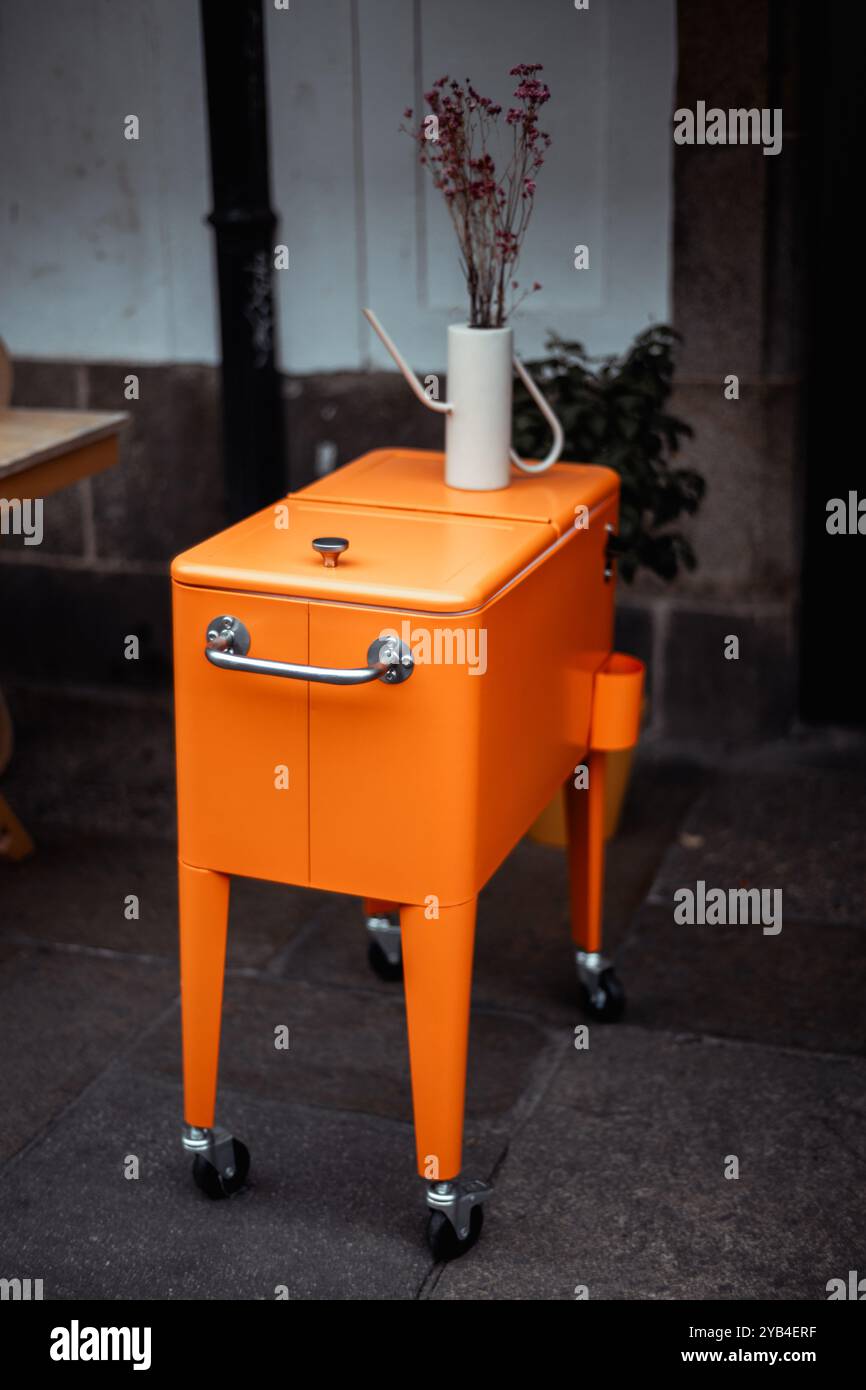 A vertical shot of an orange, modern metal cooler on wheels with a ...