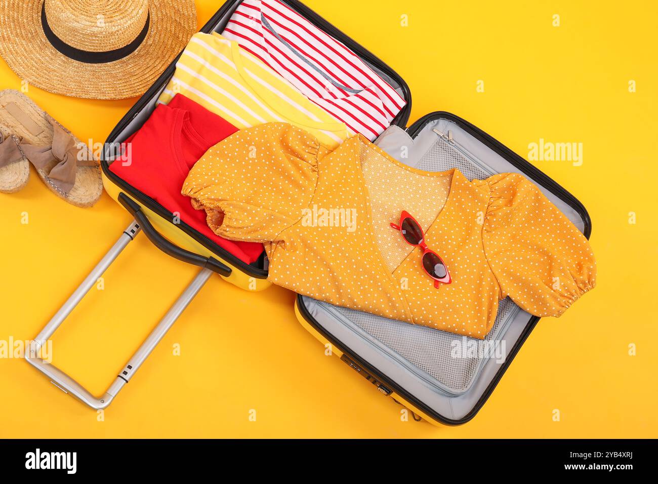 Open suitcase with clothes, wicker hat and shoes on yellow background ...
