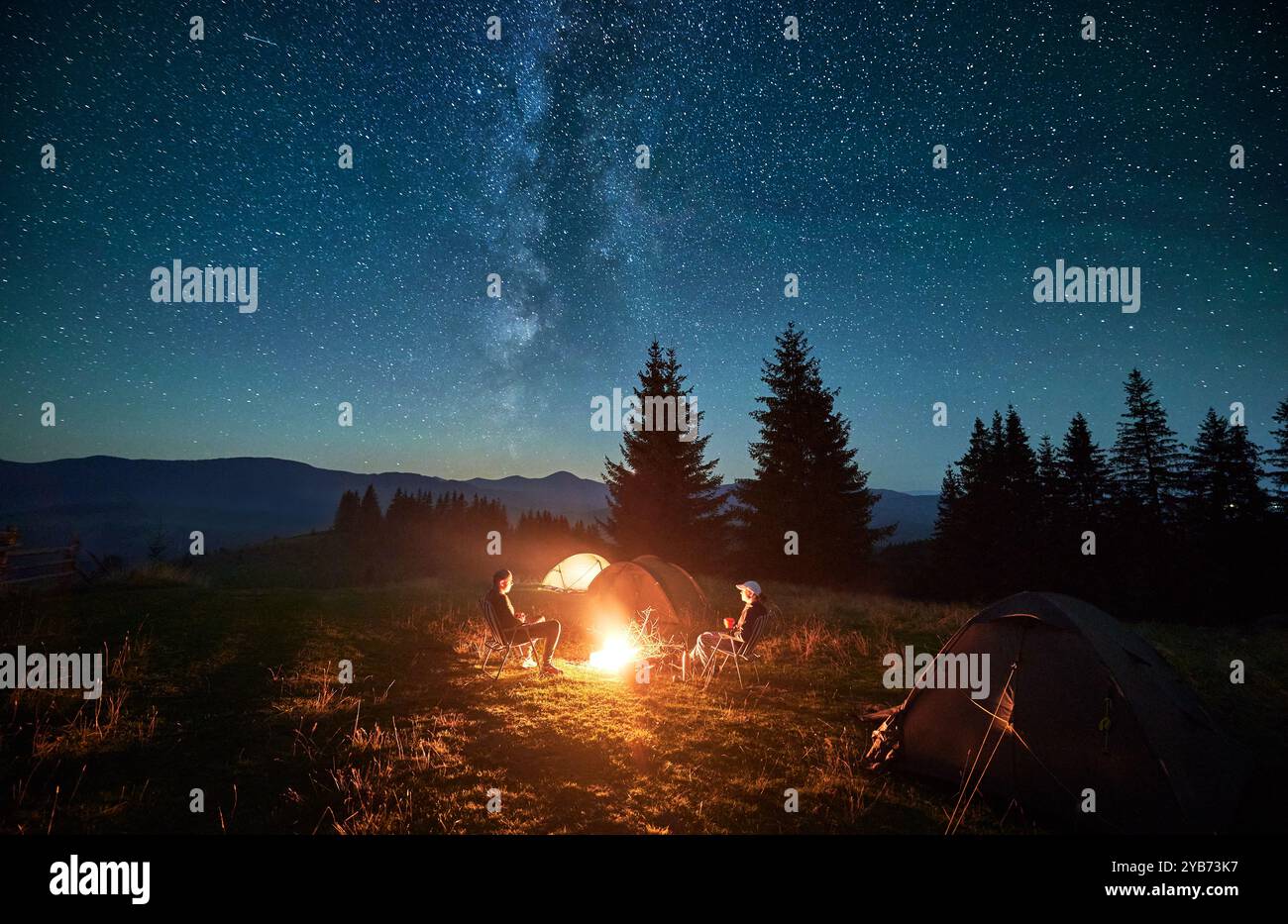 Silhouette of couple hikers resting by campfire under star-filled sky, near illuminated tents at ...