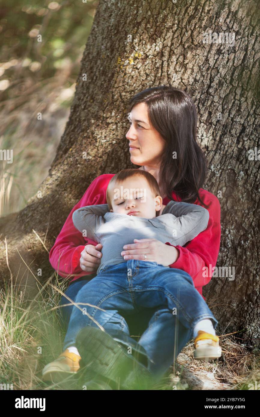 A woman is holding a sleeping baby under a tree. The scene is peaceful ...