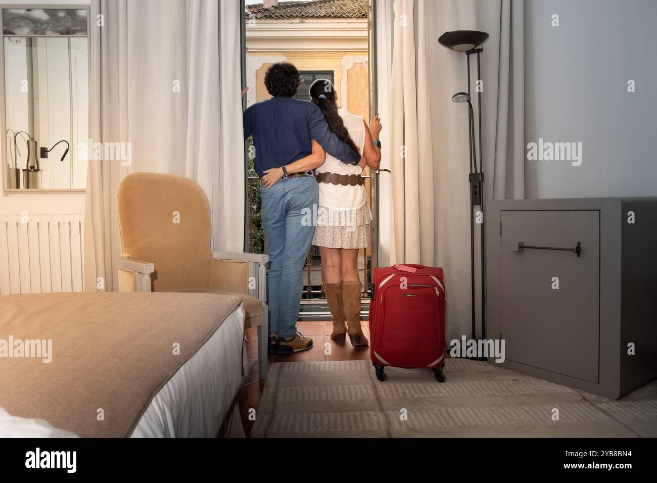 A couple facing away with a red suitcase in a hotel room with the ...