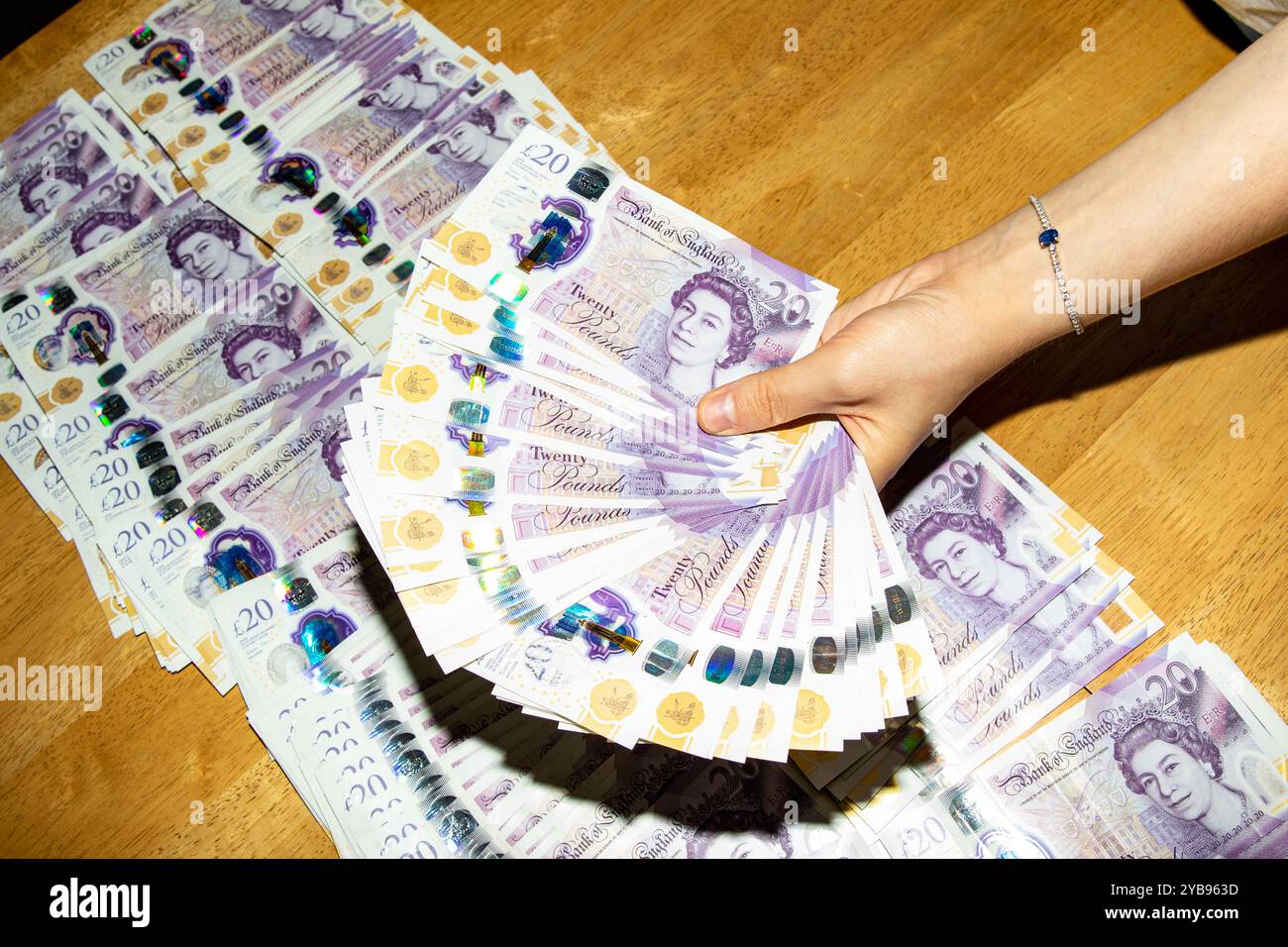 A hand holding a fan of twenty-pound banknotes, with stacks of more ...