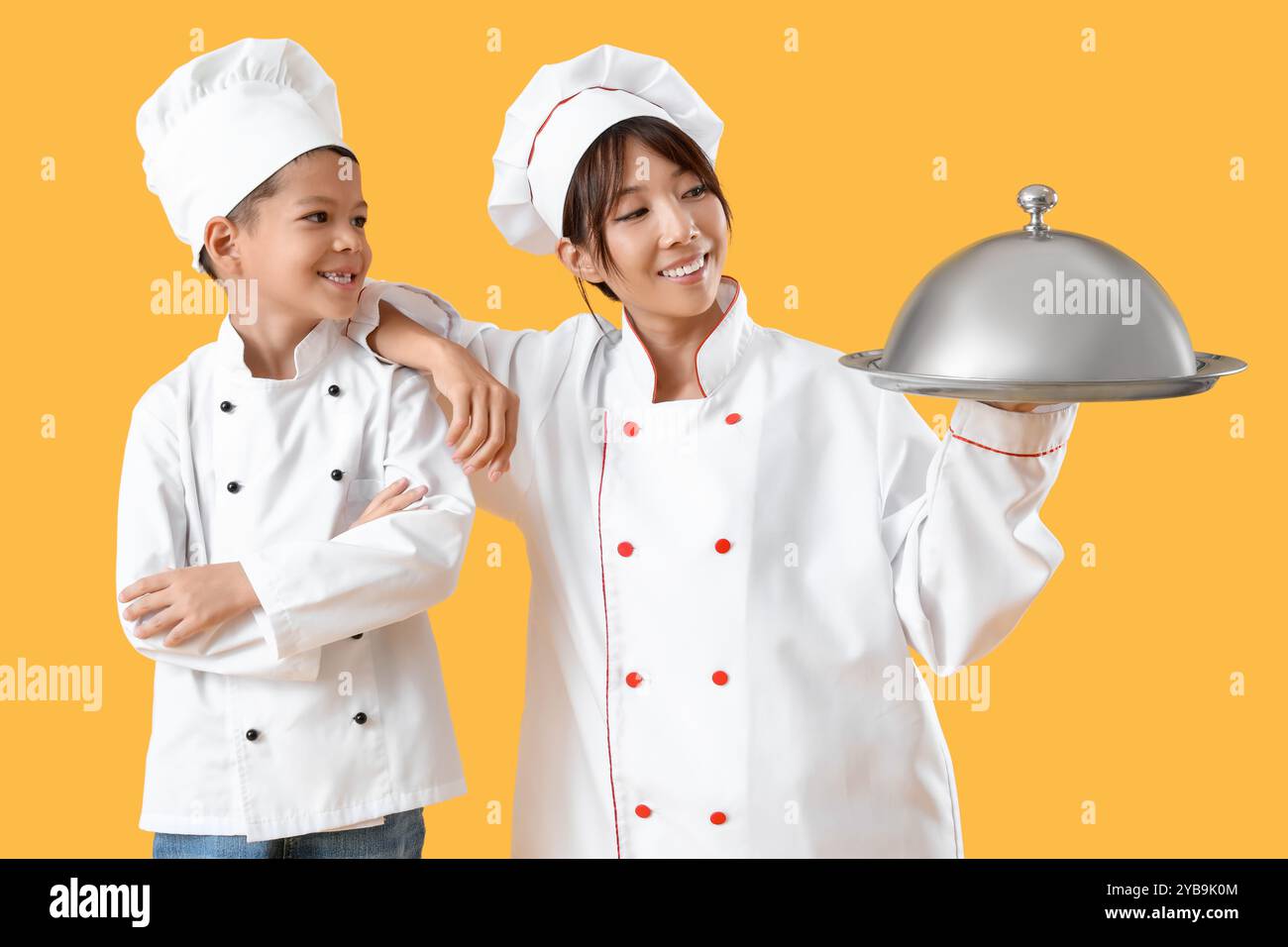Female Asian chef with cloche and her little son on yellow background ...