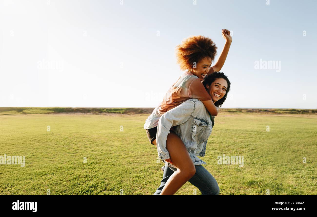 Two young women enjoying a victorious piggyback ride in a sunny field ...