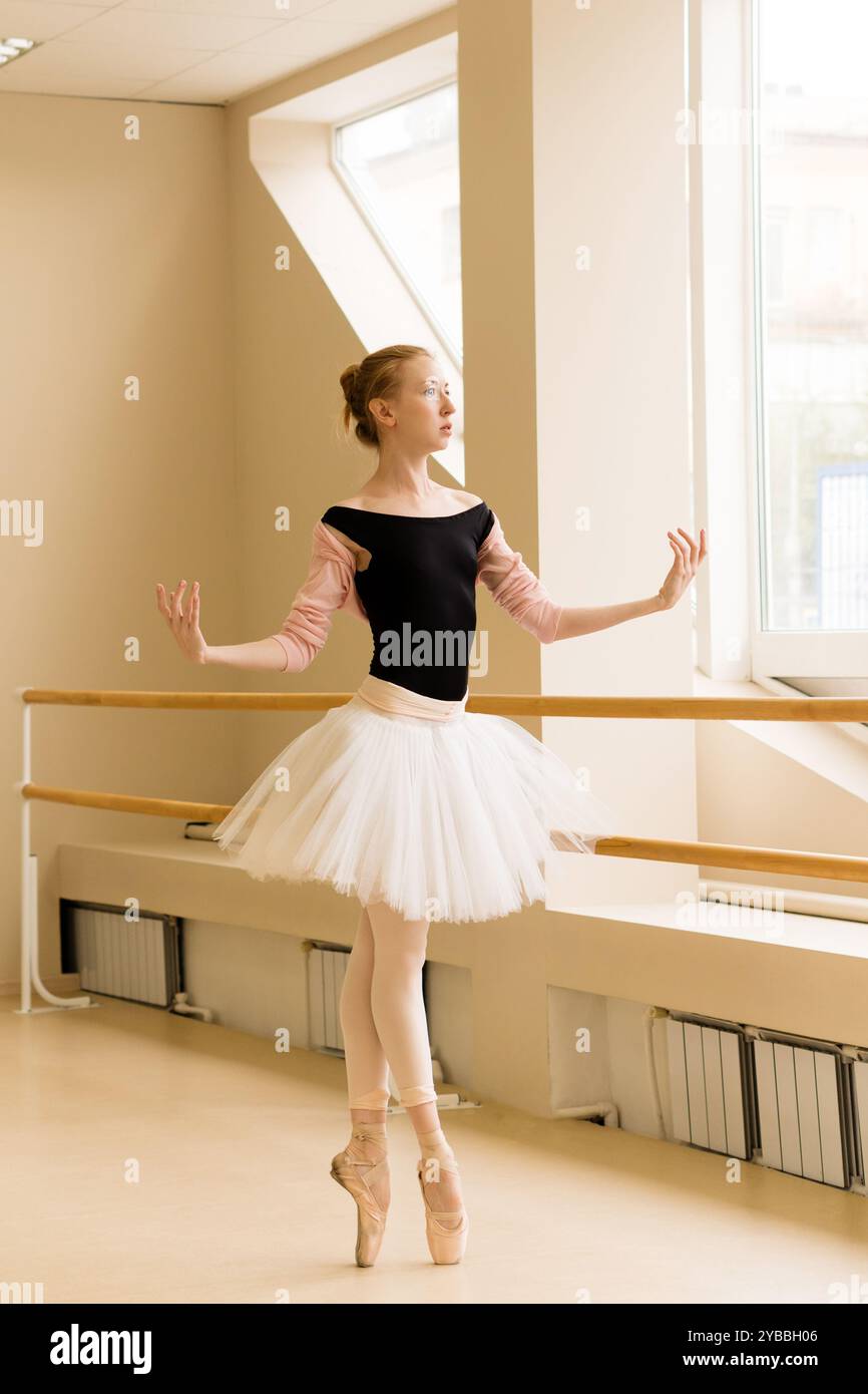 A focused ballerina practices on pointe, standing near the ballet barre ...