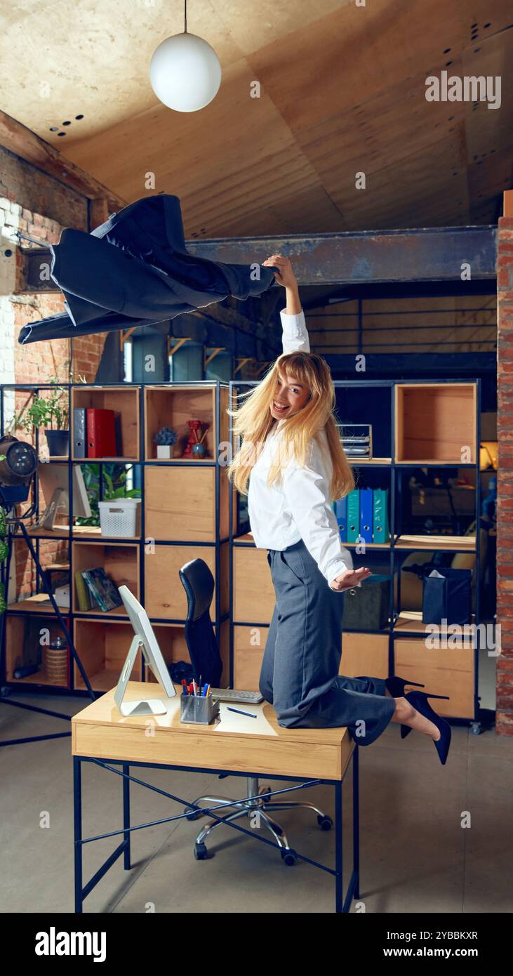 Emotional woman dancing her desk, throwing her jacket into air, smiling ...