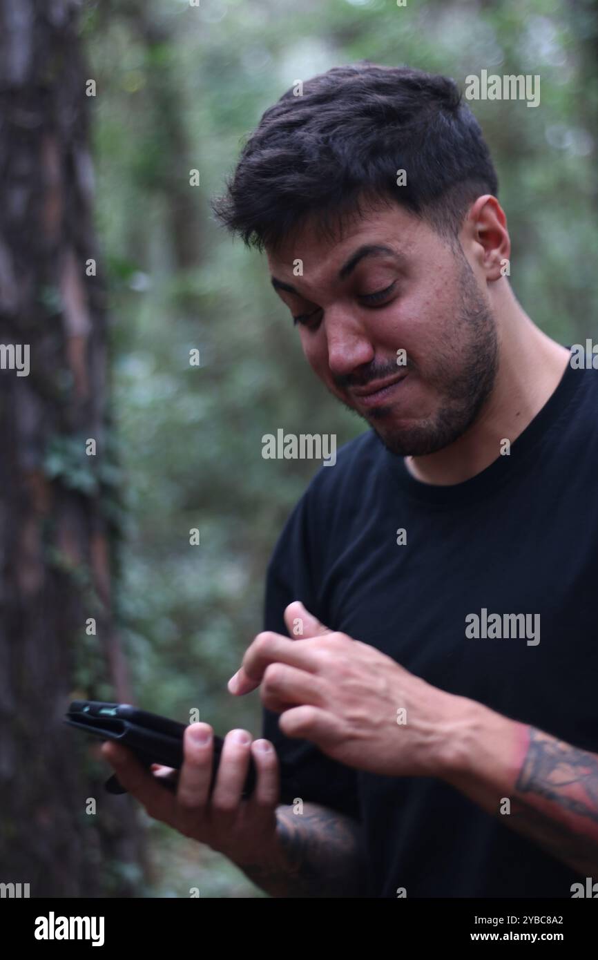 A young man stands in a peaceful forest, staring at his phone with a ...