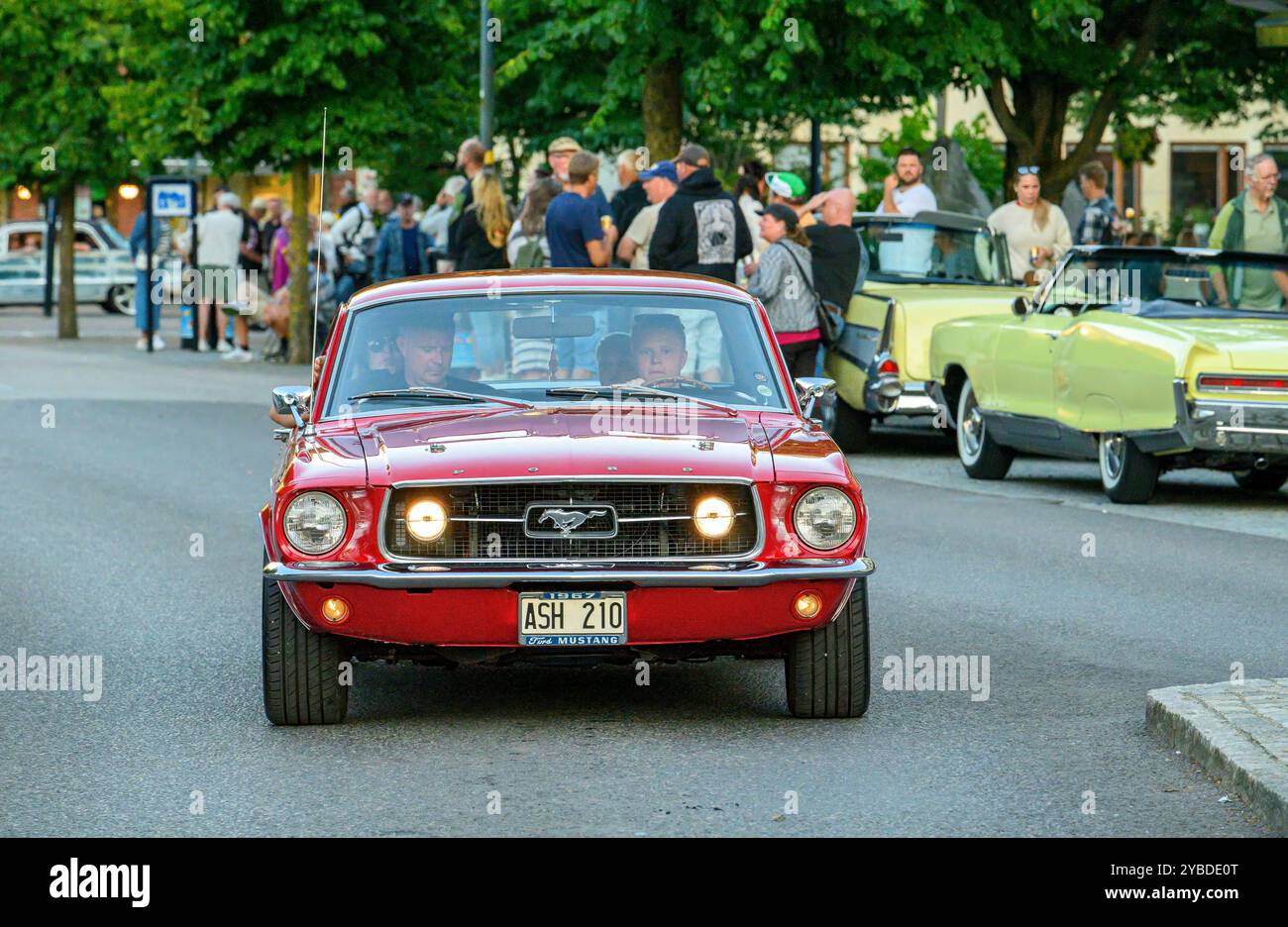A red Mustang leads a parade of classic cars, with a crowd of ...
