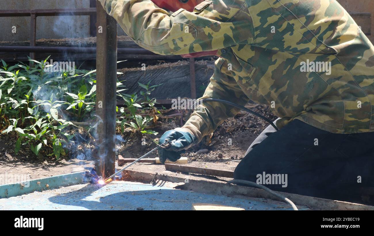 welder in green jacket and red mask welds metal beam to iron base ...