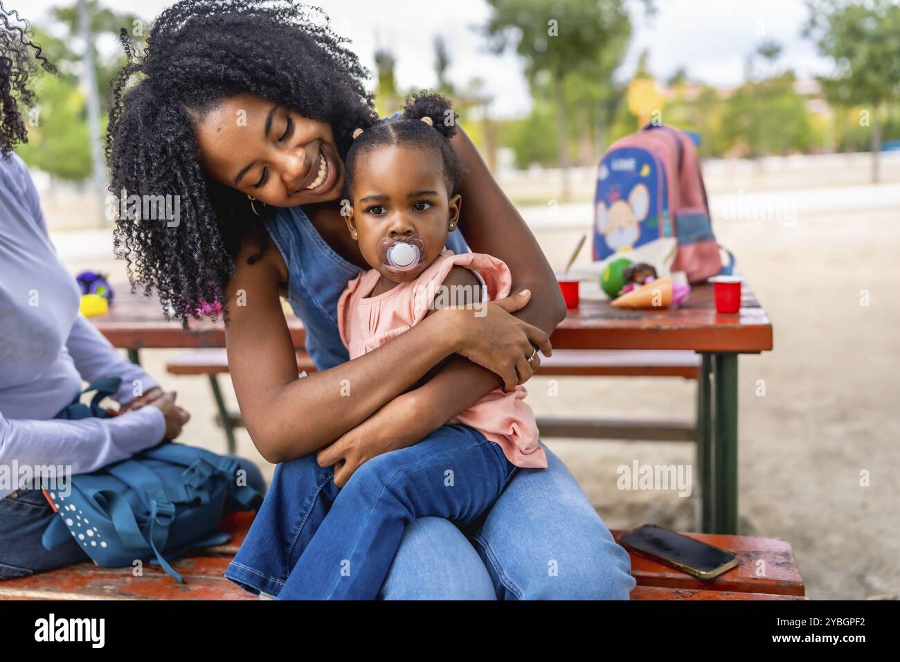 Happy african mother sitting on picnic table embracing a baby girl with ...