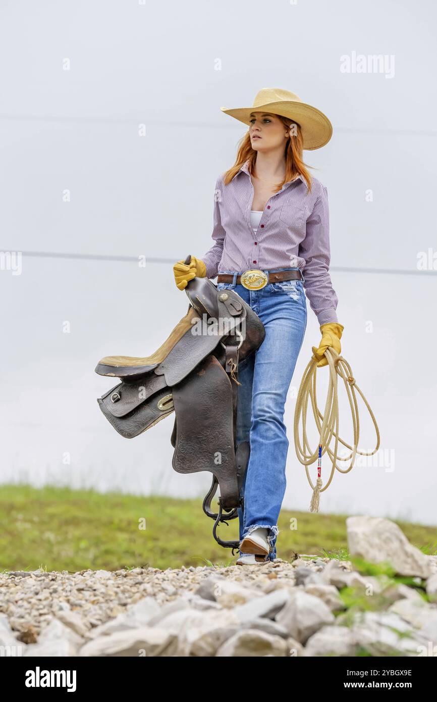 A beautiful redhead model poses in a country setting wearing cowgirl attire and holding a saddle ...