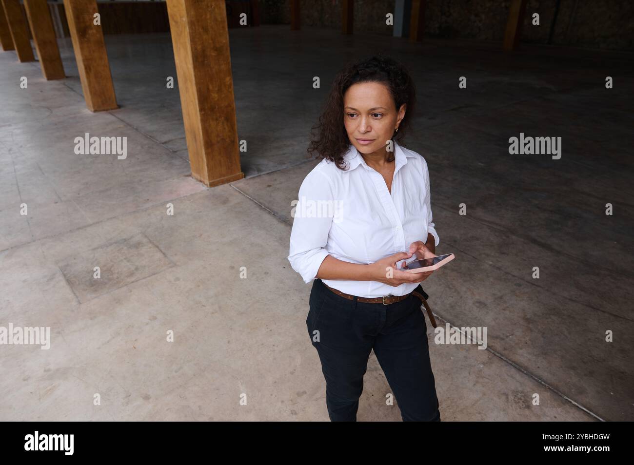 A confident professional woman stands thoughtfully holding a smartphone ...