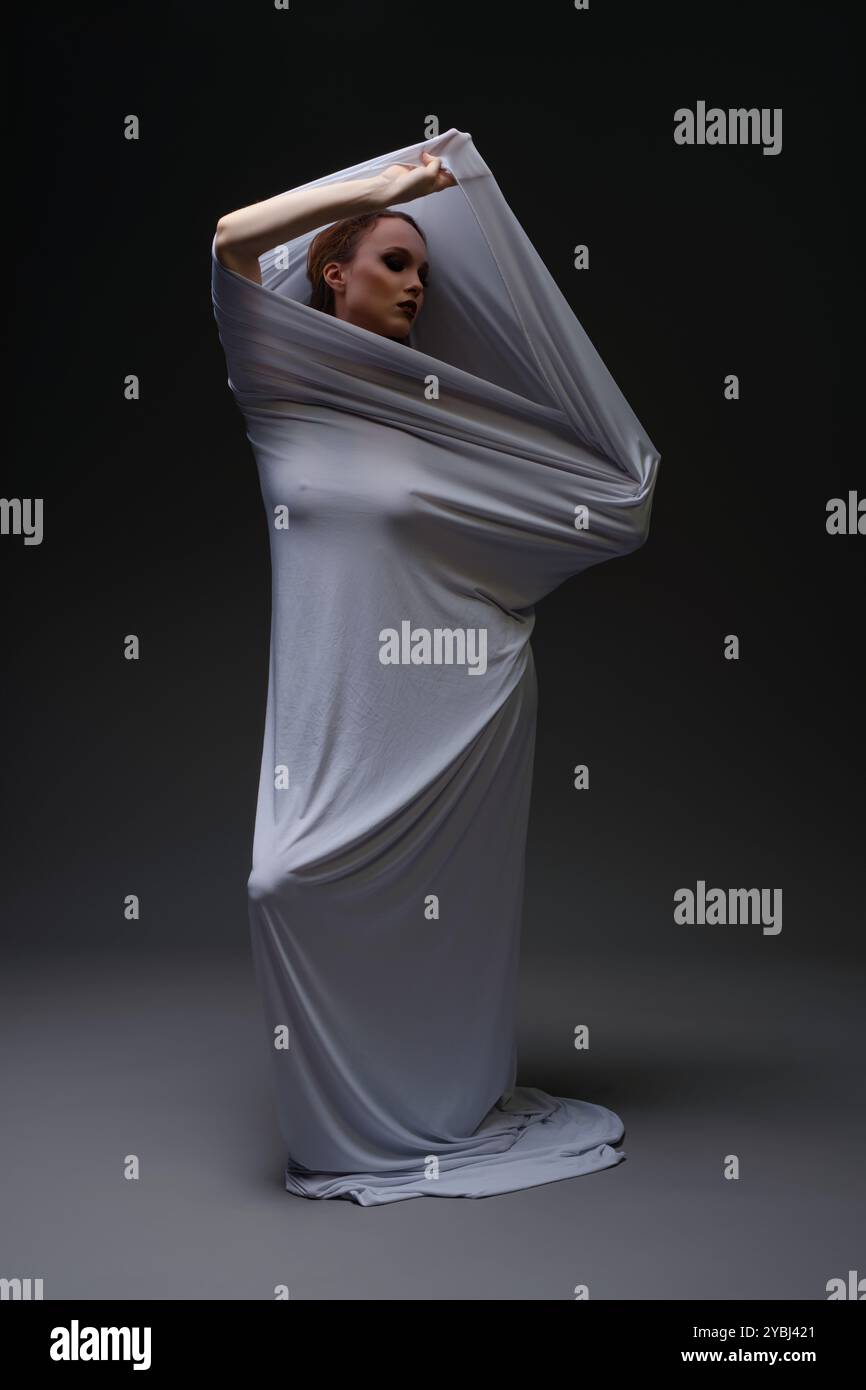A model gracefully poses in a draped white fabric in a dark studio ...