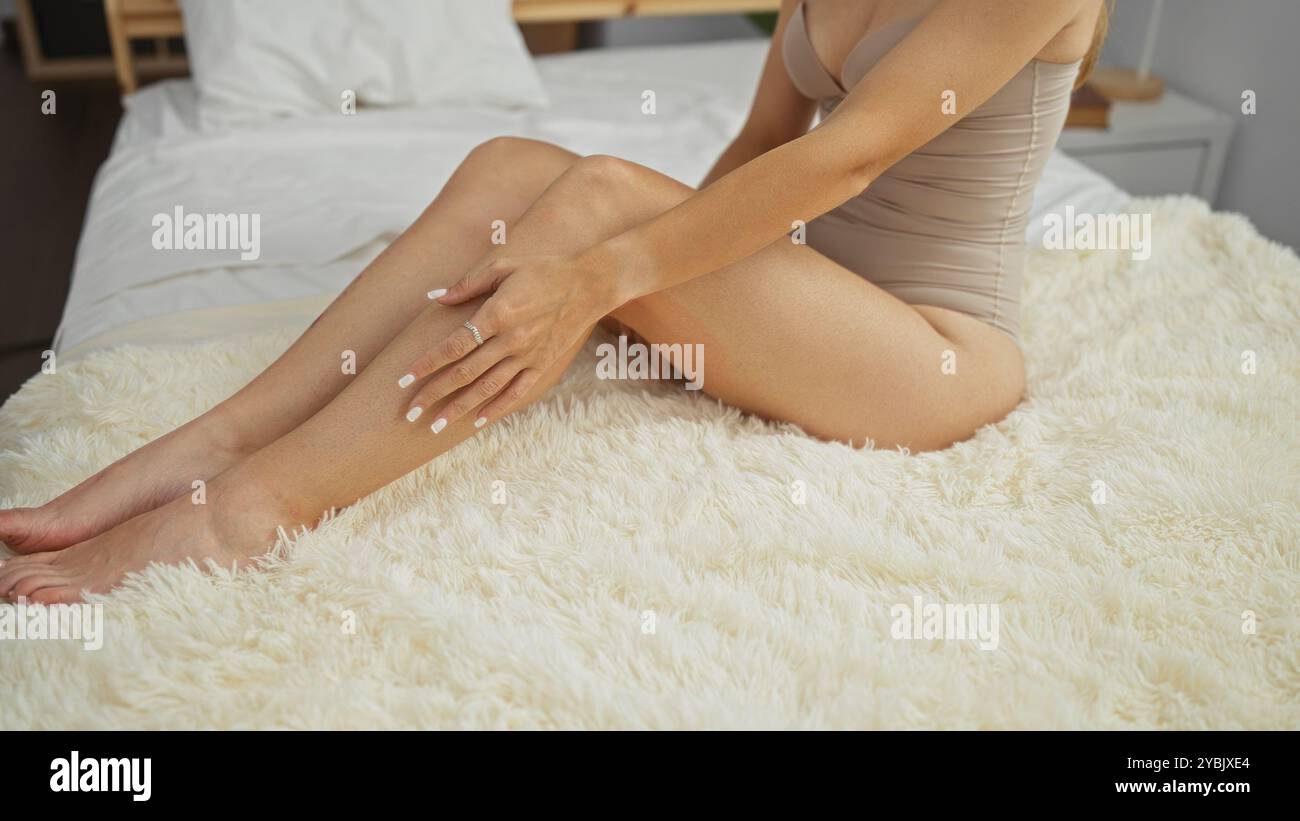 Woman sitting on bed in bedroom touching legs with hands in cozy indoor home interior Stock ...