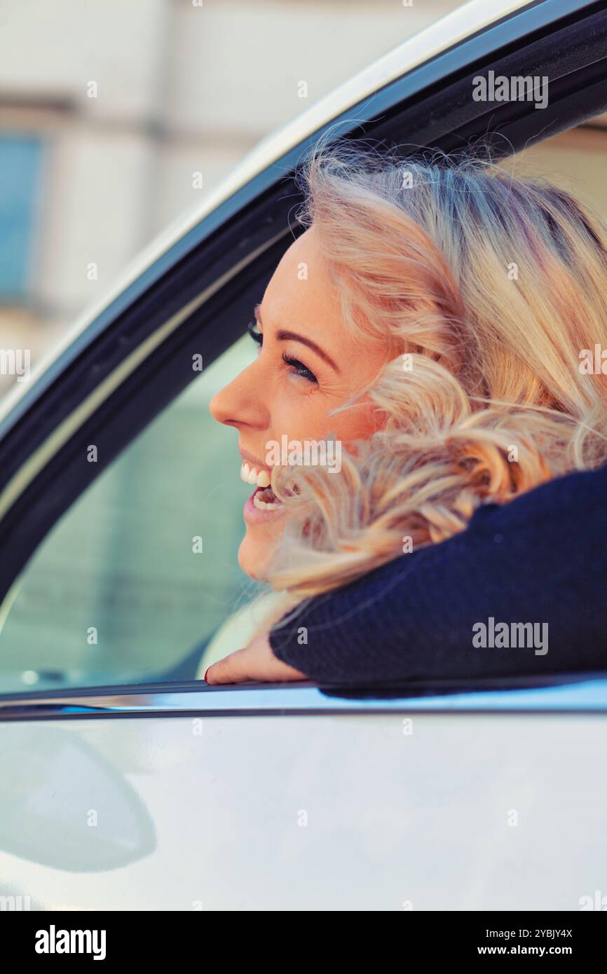 Excited blonde woman leaning out of car window on road trip, enjoying ...