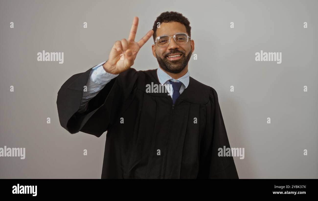 Young hispanic man smiling wearing graduation gown giving peace sign ...