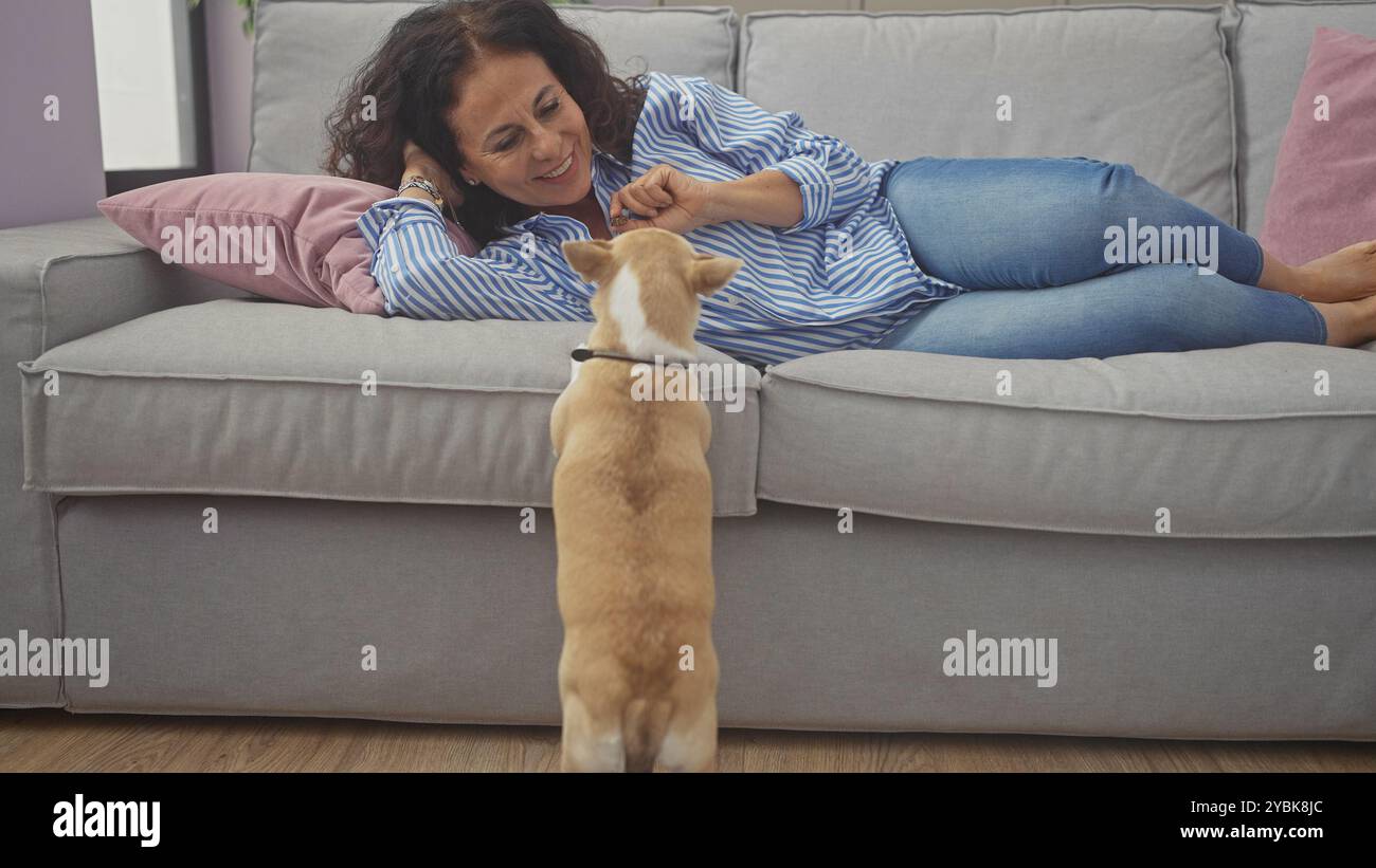 A middle-aged hispanic woman relaxes on her sofa, interacting playfully with her pet chihuahua ...