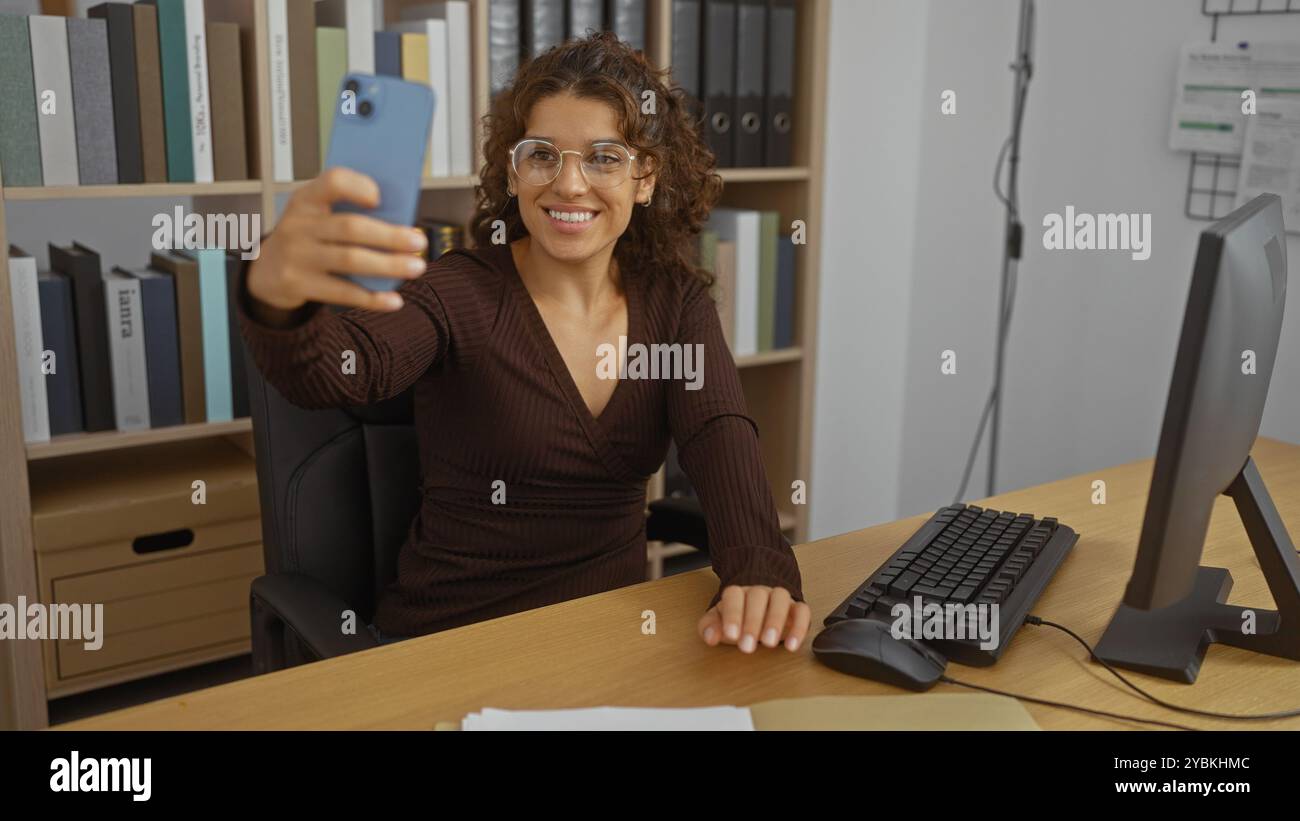 Young woman taking selfie with smartphone at office desk with computer ...