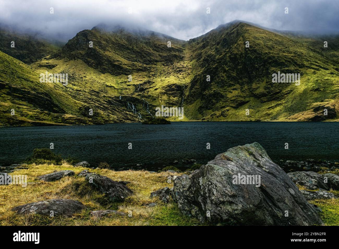 A rocky shore slopes down to a still, dark water lake, with two large ...