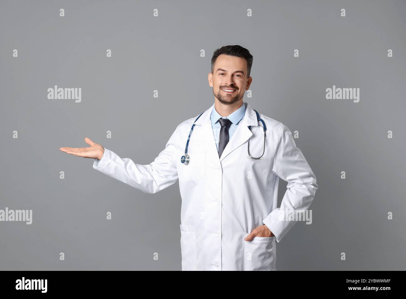 Smiling doctor with stethoscope pointing at something on grey ...