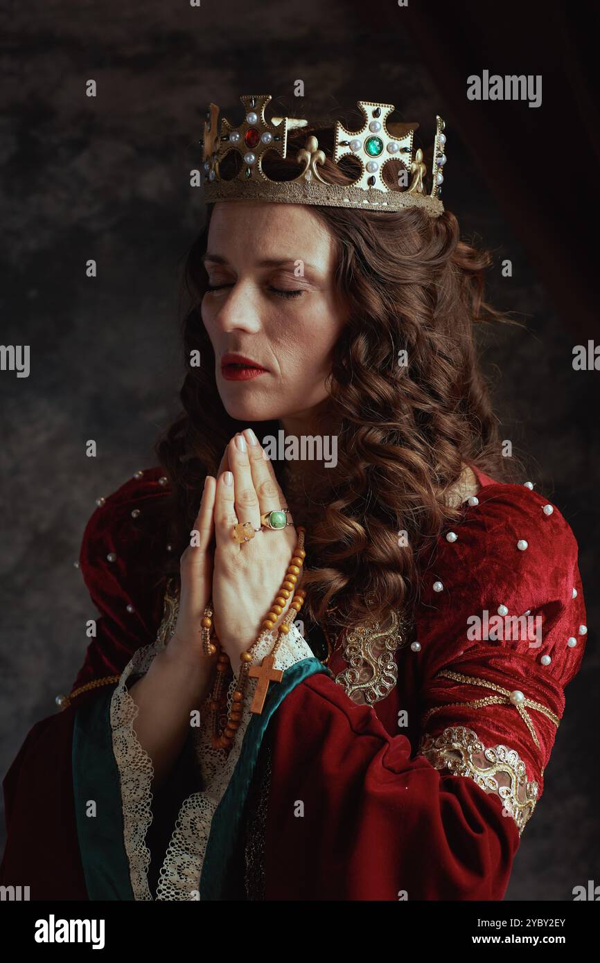 medieval queen in red dress with rosary and crown praying on dark gray ...