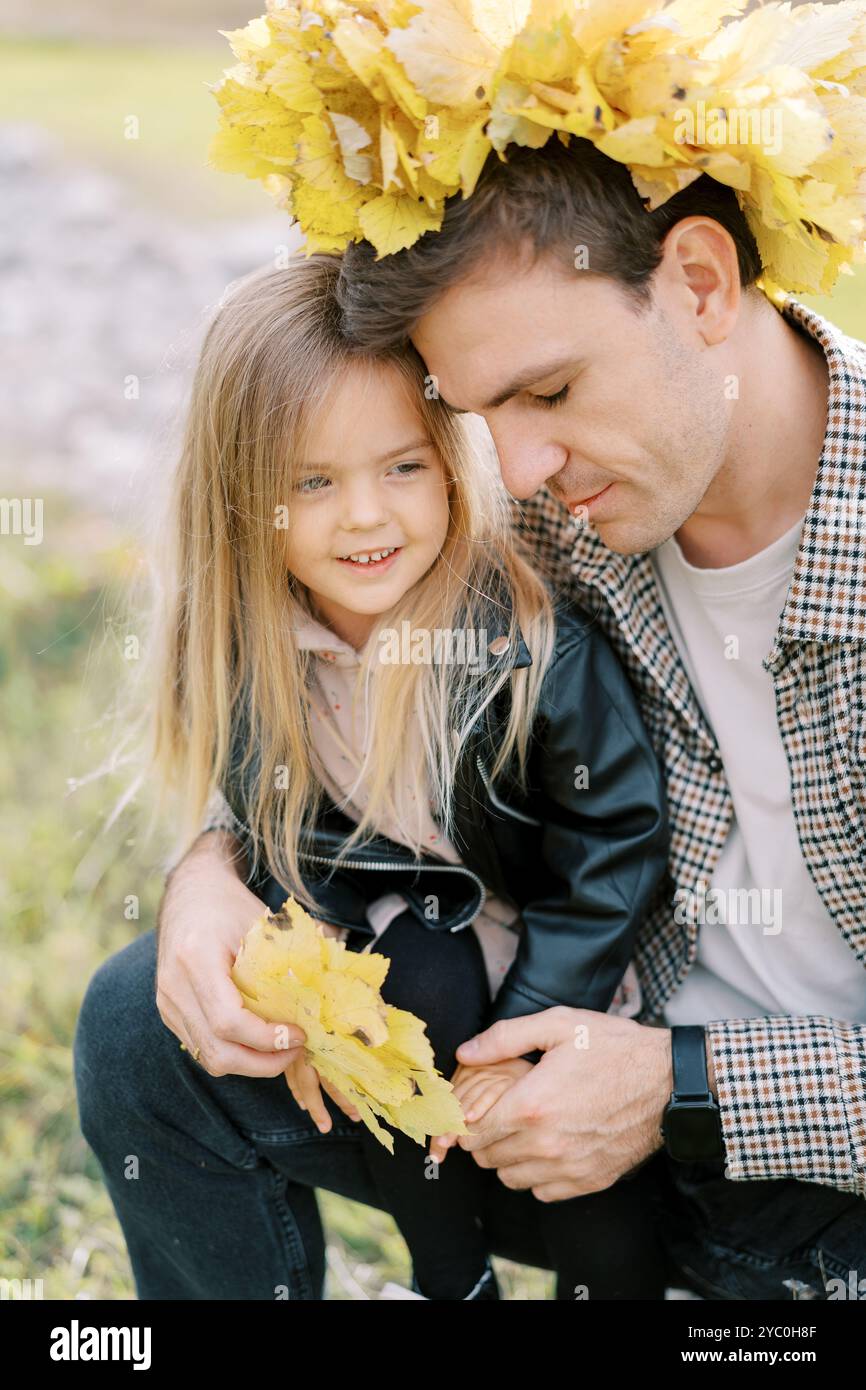 Dad in a wreath of yellow leaves hugs a little girl sitting on his lap, touching foreheads Stock ...
