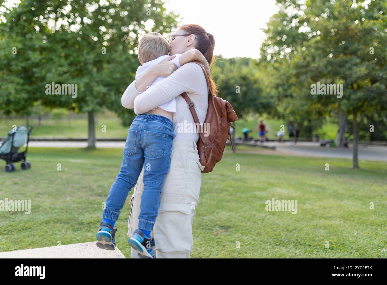 A woman is hugging a child in a park. The child is wearing a backpack ...