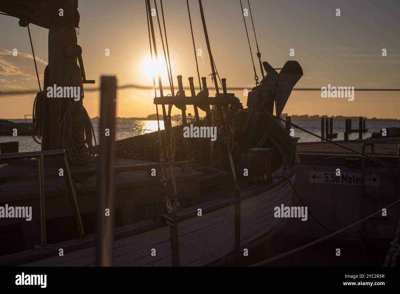 A historic wooden sailing ship docked at sunset, casting long shadows ...