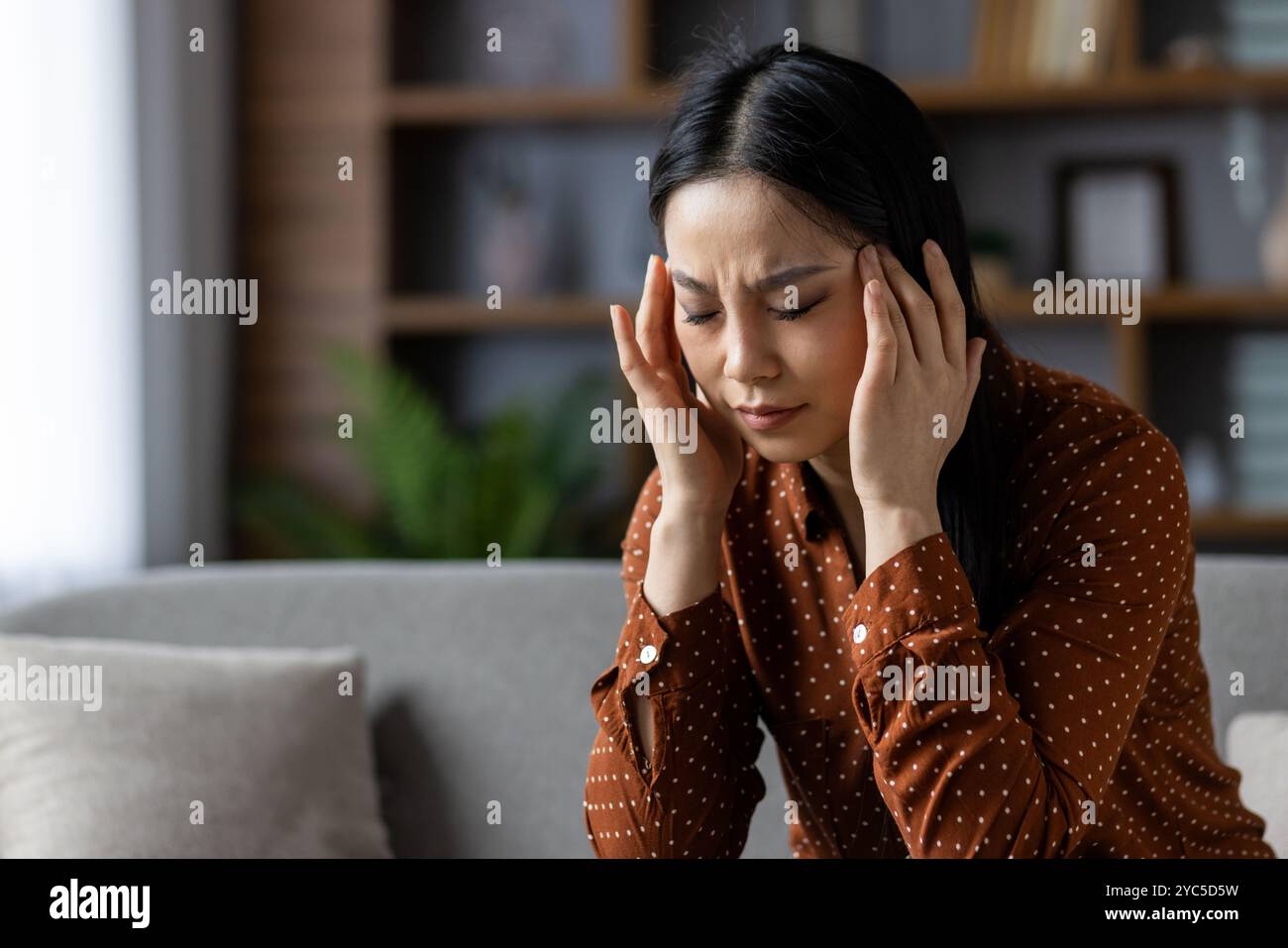 Asian woman experiencing stress. She sits on couch holding head with ...