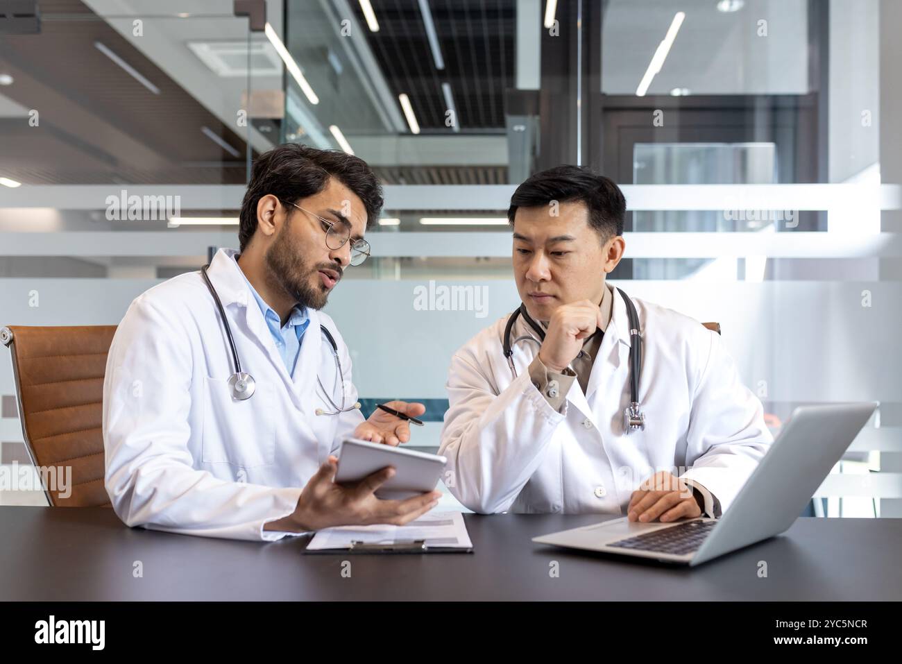 Doctors engaged in discussion over patient diagnosis in an office setting, utilizing tablet and ...