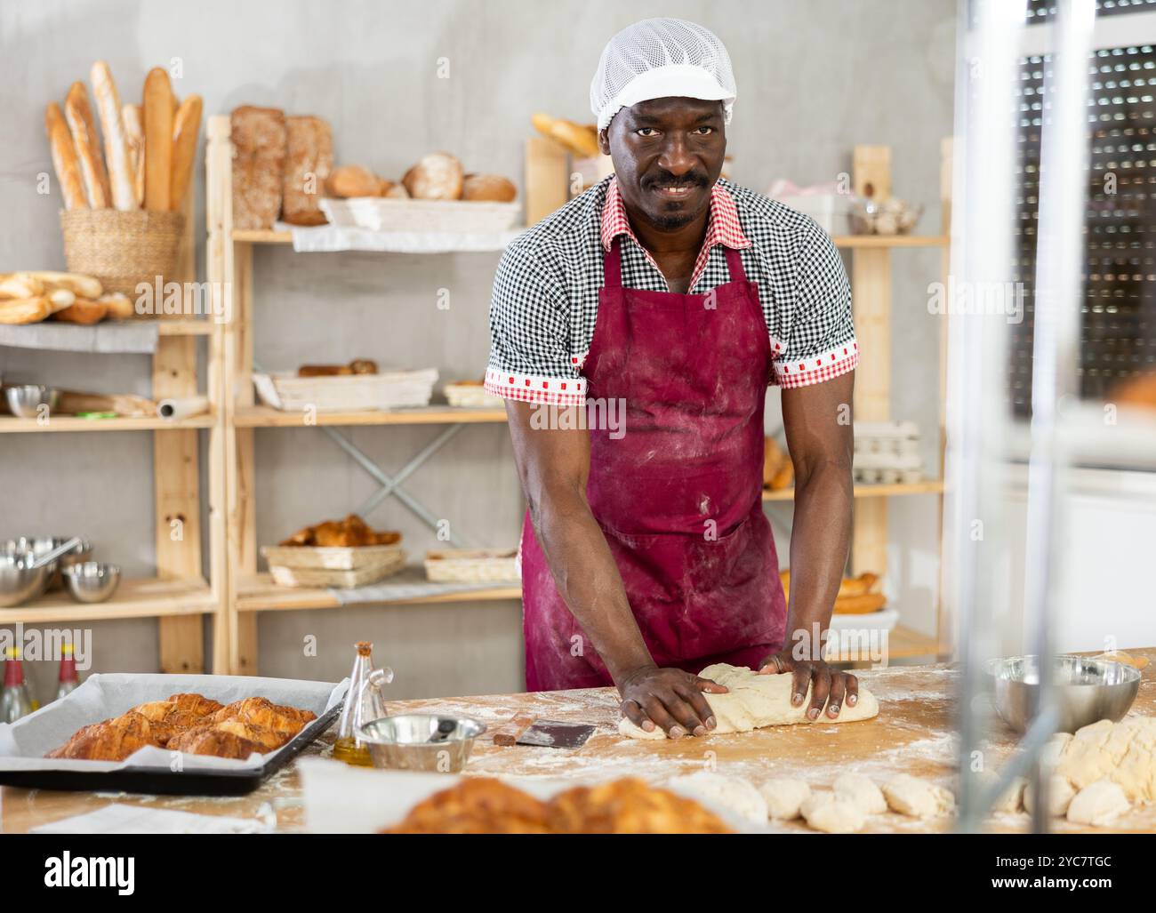 Confident male baker stands at his work bench, kneading and shaping ...