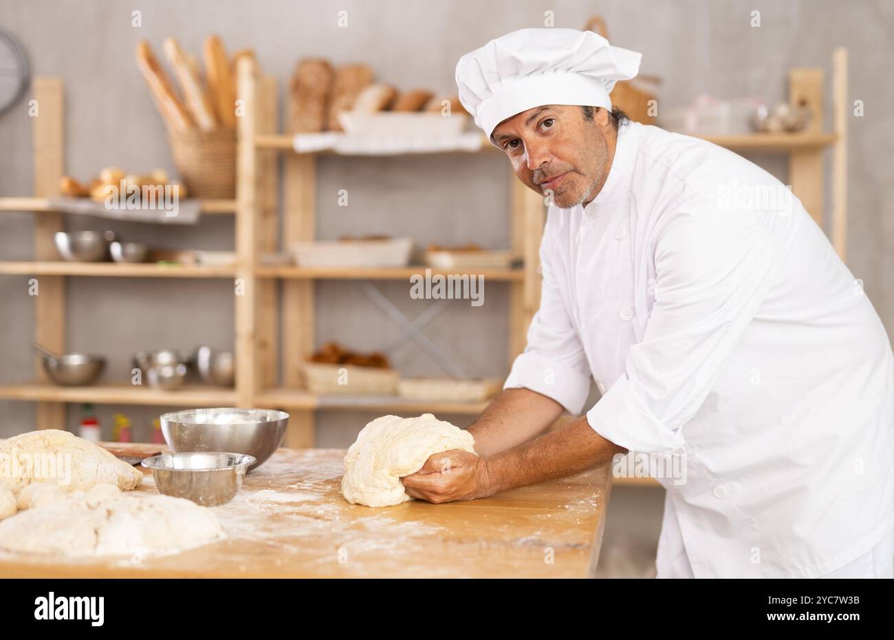 Male baker stands at work bench, kneading and shaping dough to make ...
