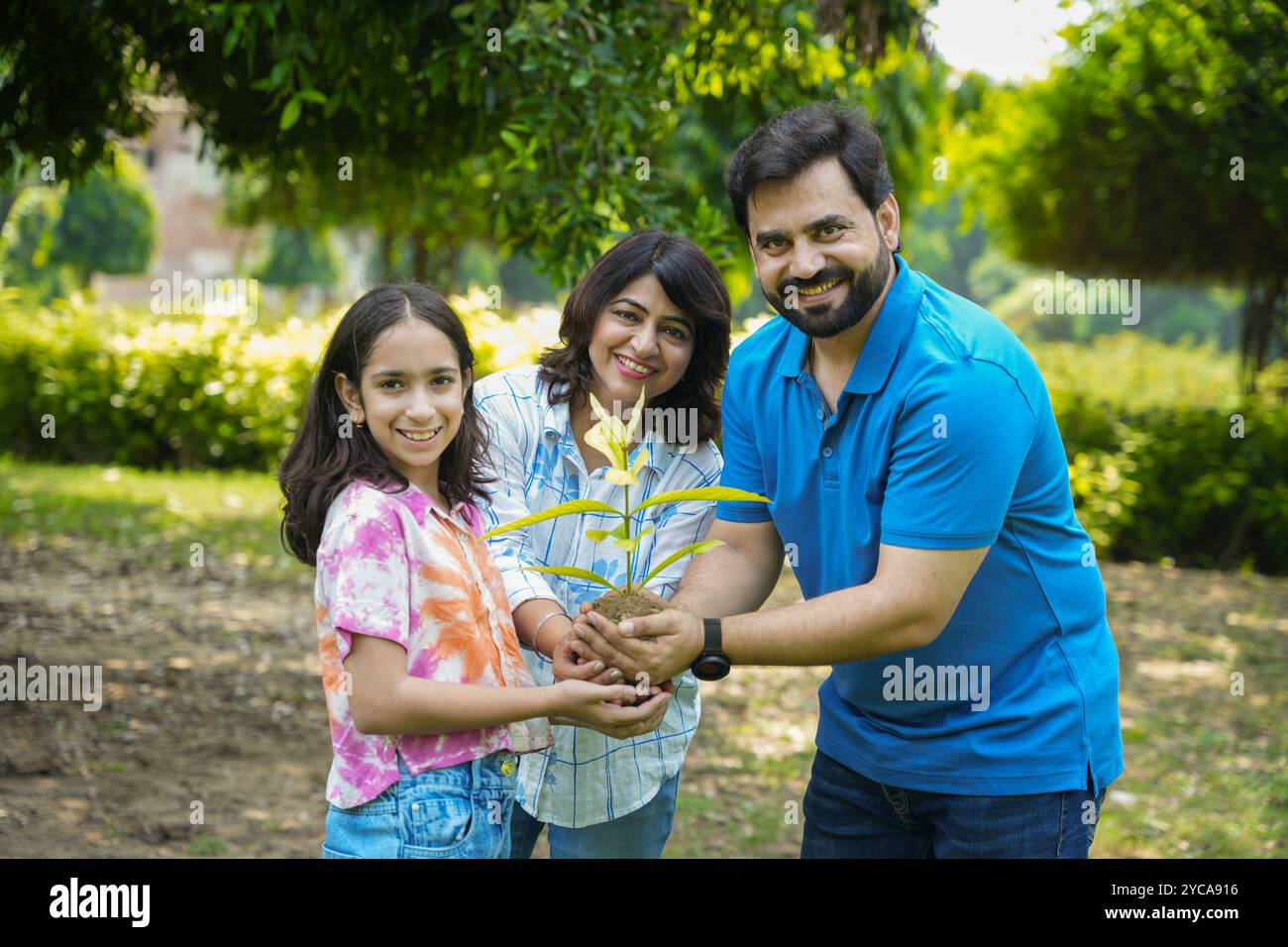 happy young indian family of three planting tree while looking at ...