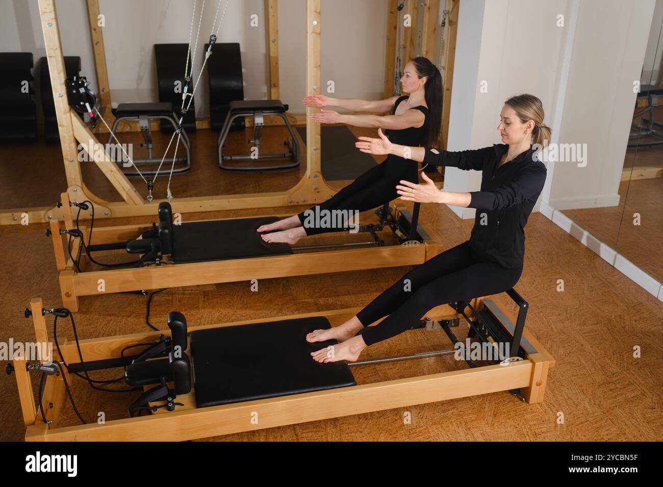 Two women sitting on a reformer exercise machine perform an exercise in ...