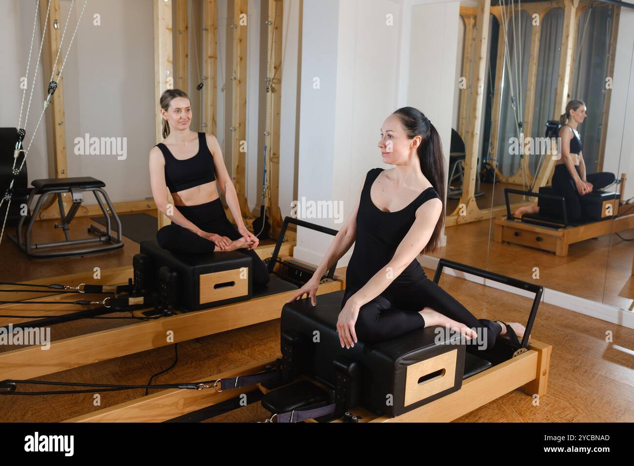 Two women sitting on a reformer exercise machine perform an exercise in ...