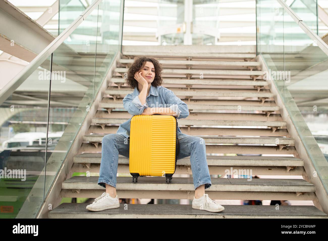 Young woman relaxing on stairs with a bright yellow suitcase in a busy ...