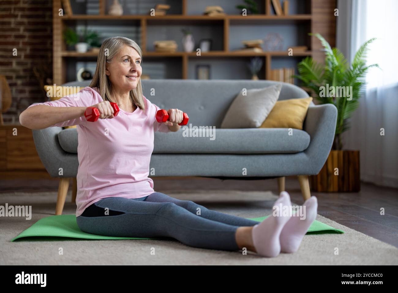 Smiling senior woman exercises with dumbbells at home, promoting active ...