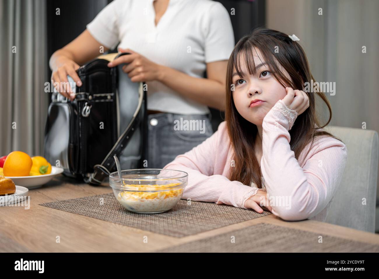 A young, stubborn Asian girl is sitting at the dining table while her ...