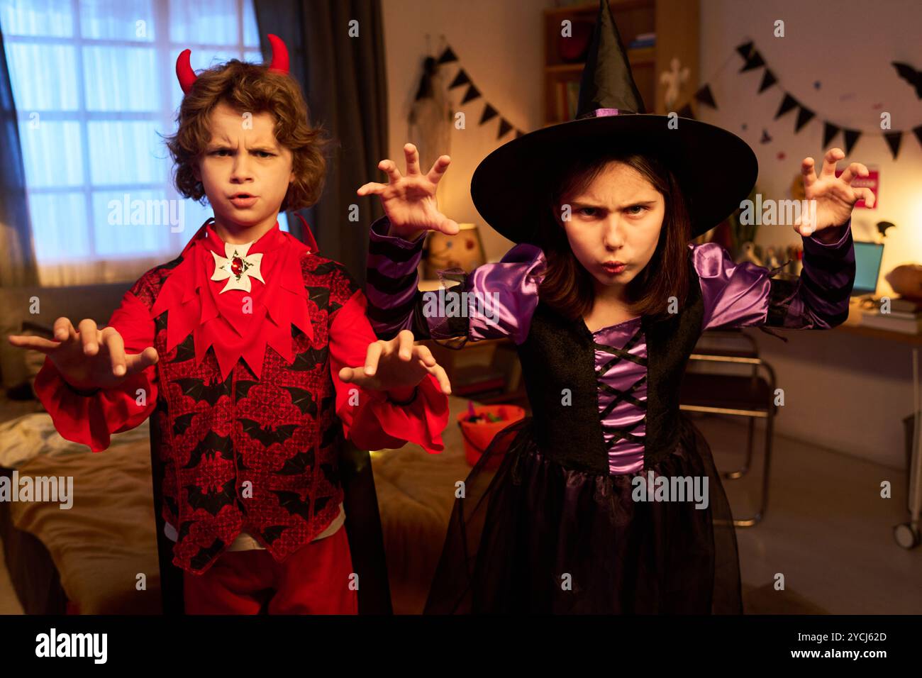 Two youthful kids in Halloween costumes of witch and demon scowling and ...