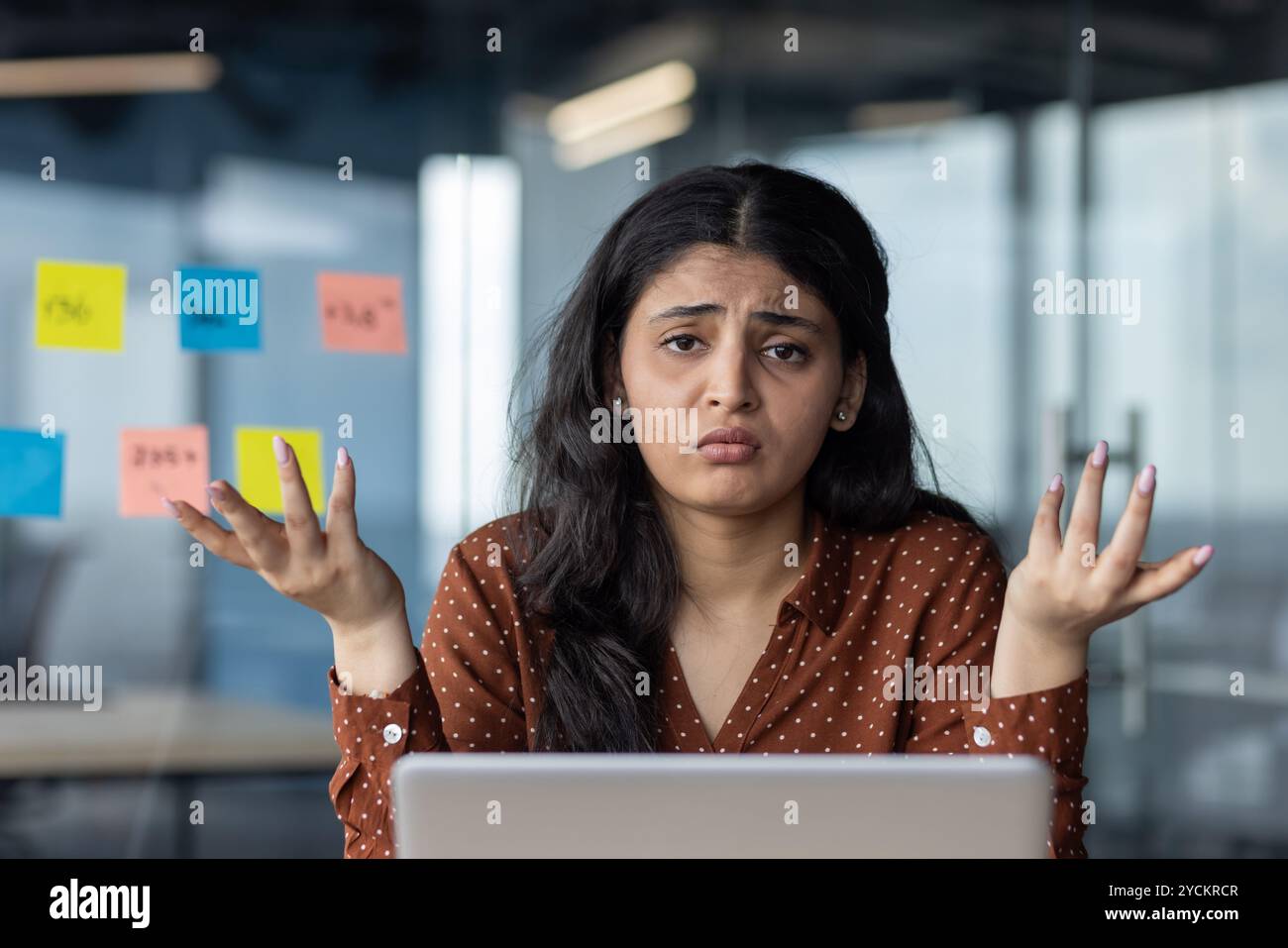 Confused Latina businesswoman shrugs baffled at desk, surrounded by ...