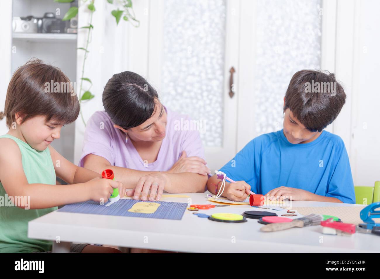A woman is helping two children make a craft. The children are wearing ...