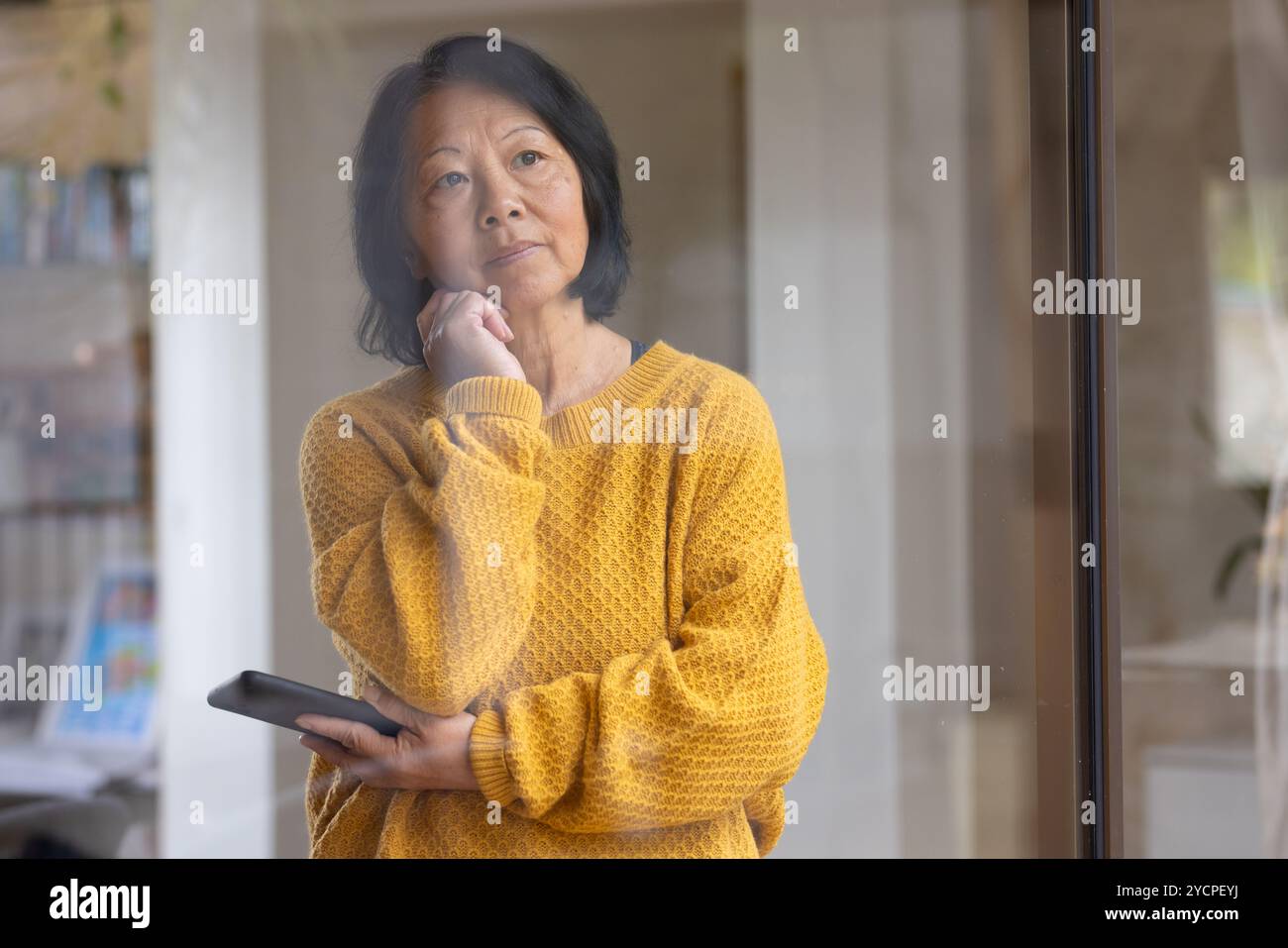 Thoughtful senior asian woman holding smartphone, gazing out window at ...