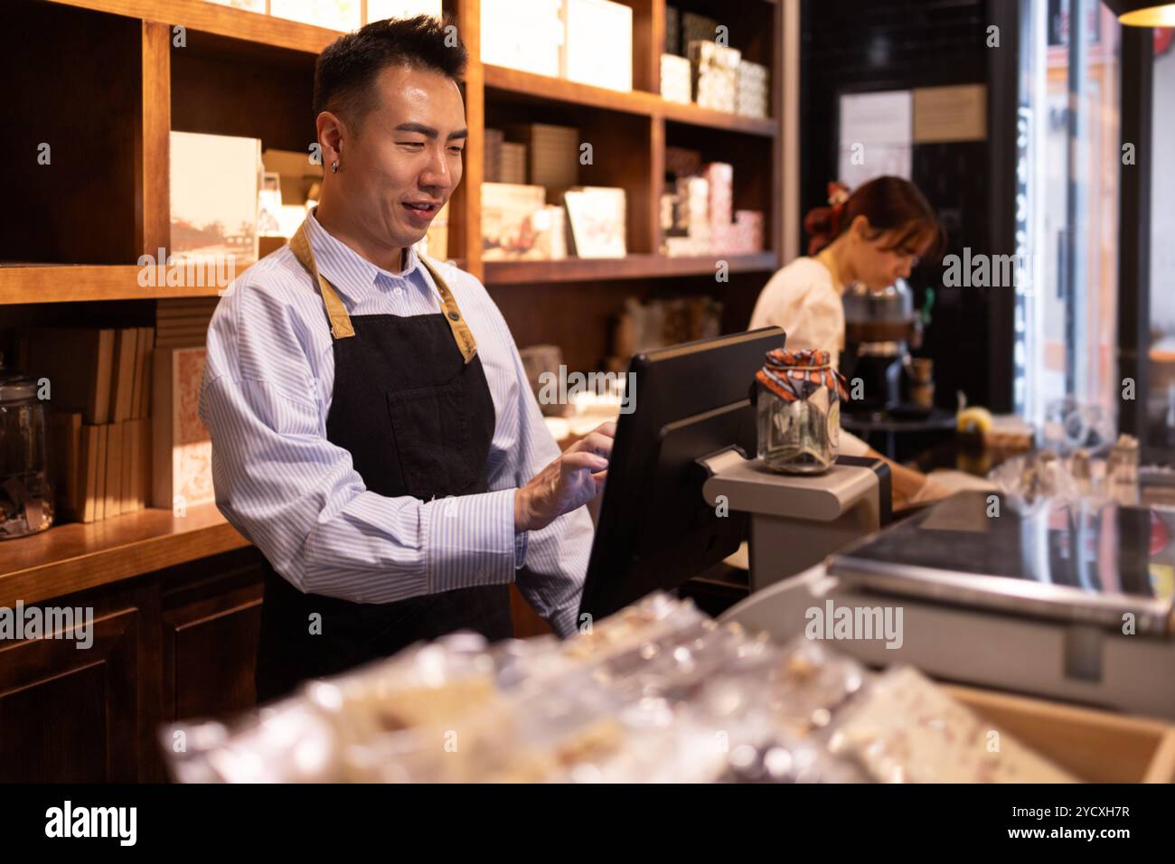 A male shopkeeper with a smiling expression operates the register at a ...